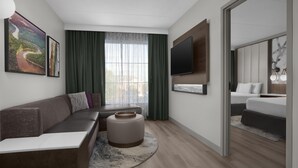 Quarto de hotel moderno com sofá em forma de L, um banquinho redondo, televisão de tela plana, cama e uma grande janela com cortinas.