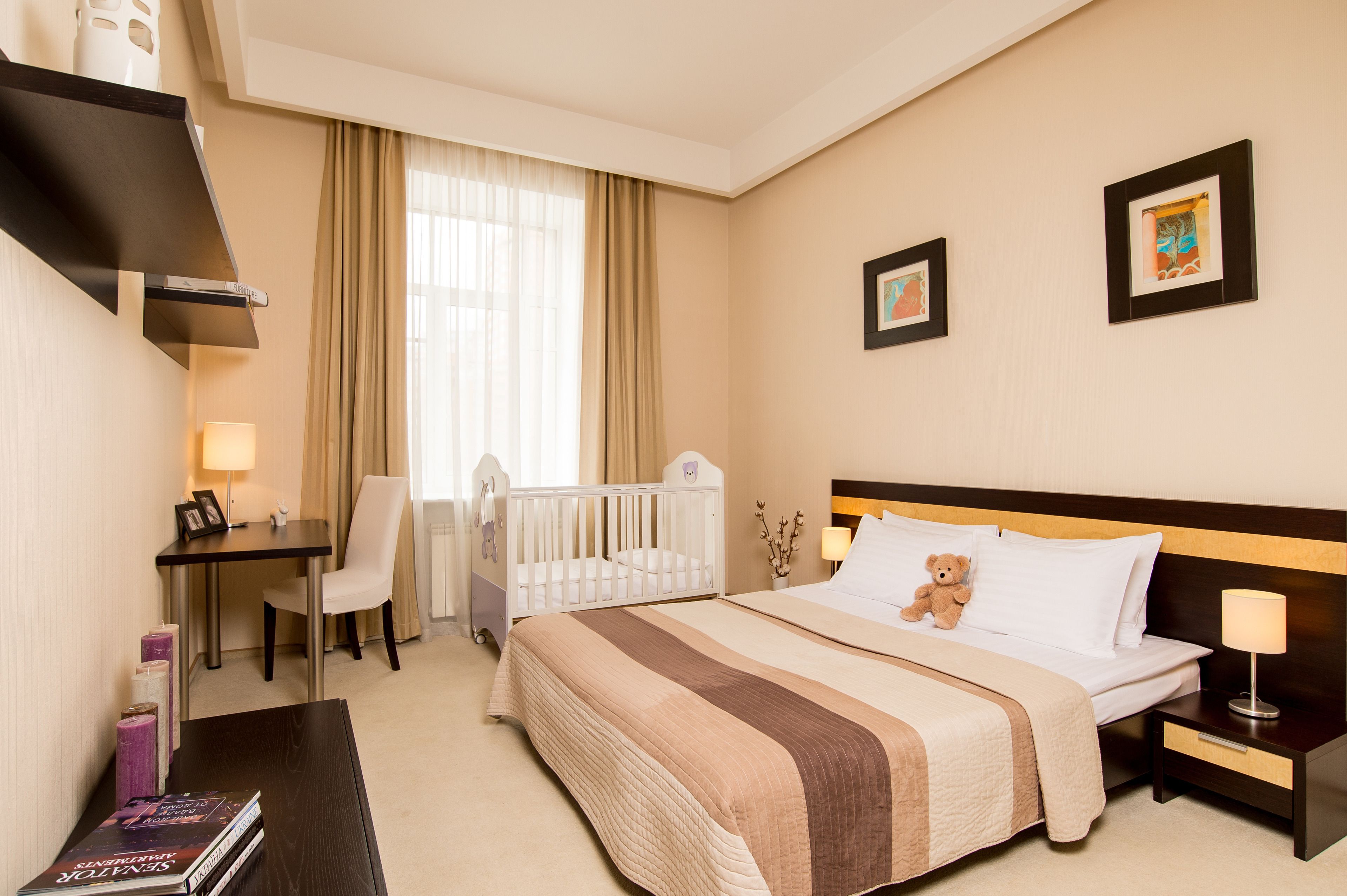 Quarto de hotel com cama, mesa de cabeceira, escrivaninha, cadeira, berço e obras de arte nas paredes.