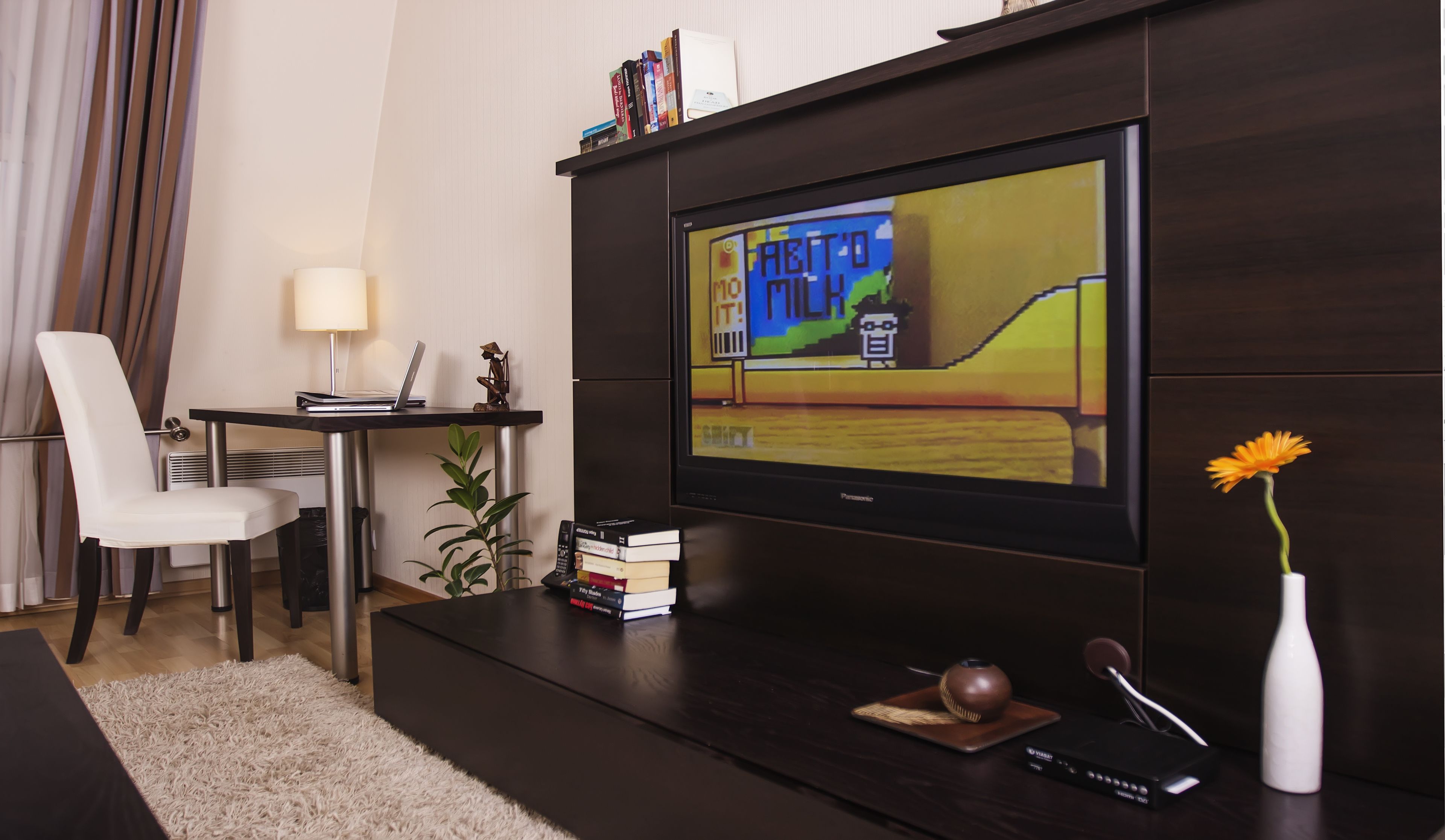 Quarto de hotel com televisão de tela plana, uma cadeira branca, uma mesinha com abajur, uma planta em vaso e um vaso com flor.