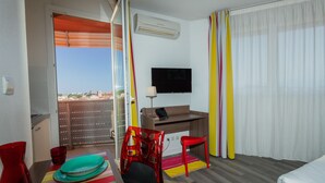 Quarto de hotel com varanda, uma escrivaninha com um laptop, televisão, cama e uma cadeira vermelha.