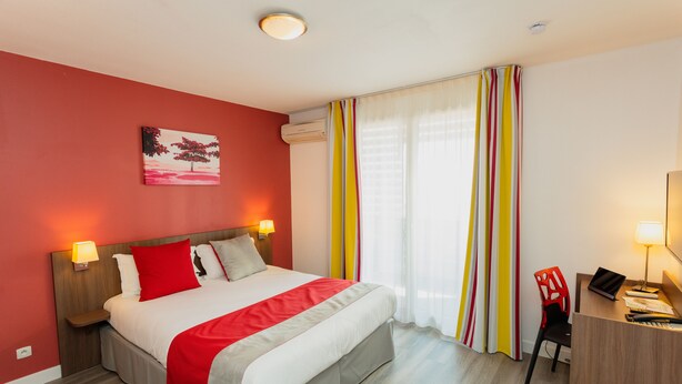 Quarto de hotel com cama, luminárias de cabeceira, uma escrivaninha com cadeira e uma janela com cortinas.