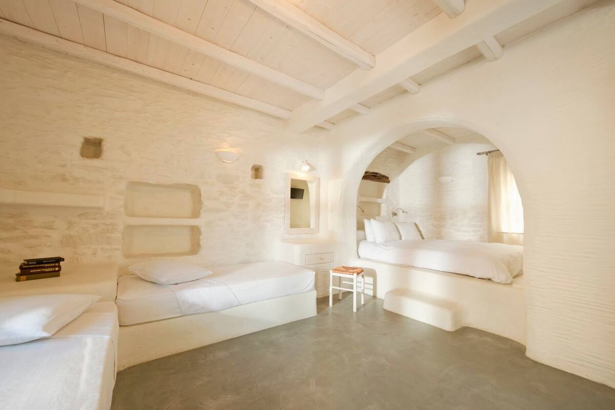 Quarto minimalista com paredes brancas, parede de destaque em pedra, um banco embutido e um pequeno banquinho.
