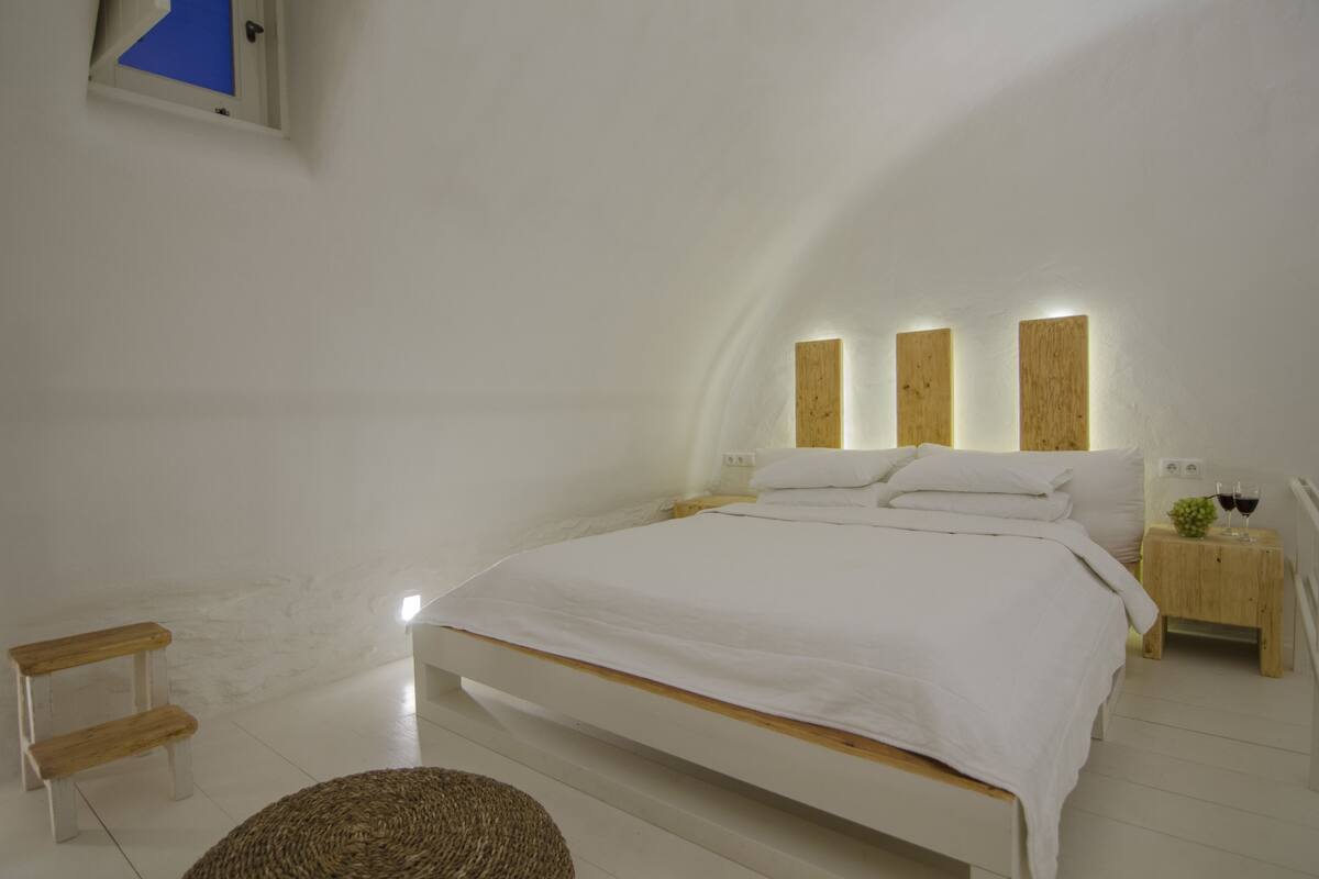 Quarto moderno com uma cama grande, mesa de cabeceira com abajur e um banquinho pequeno.