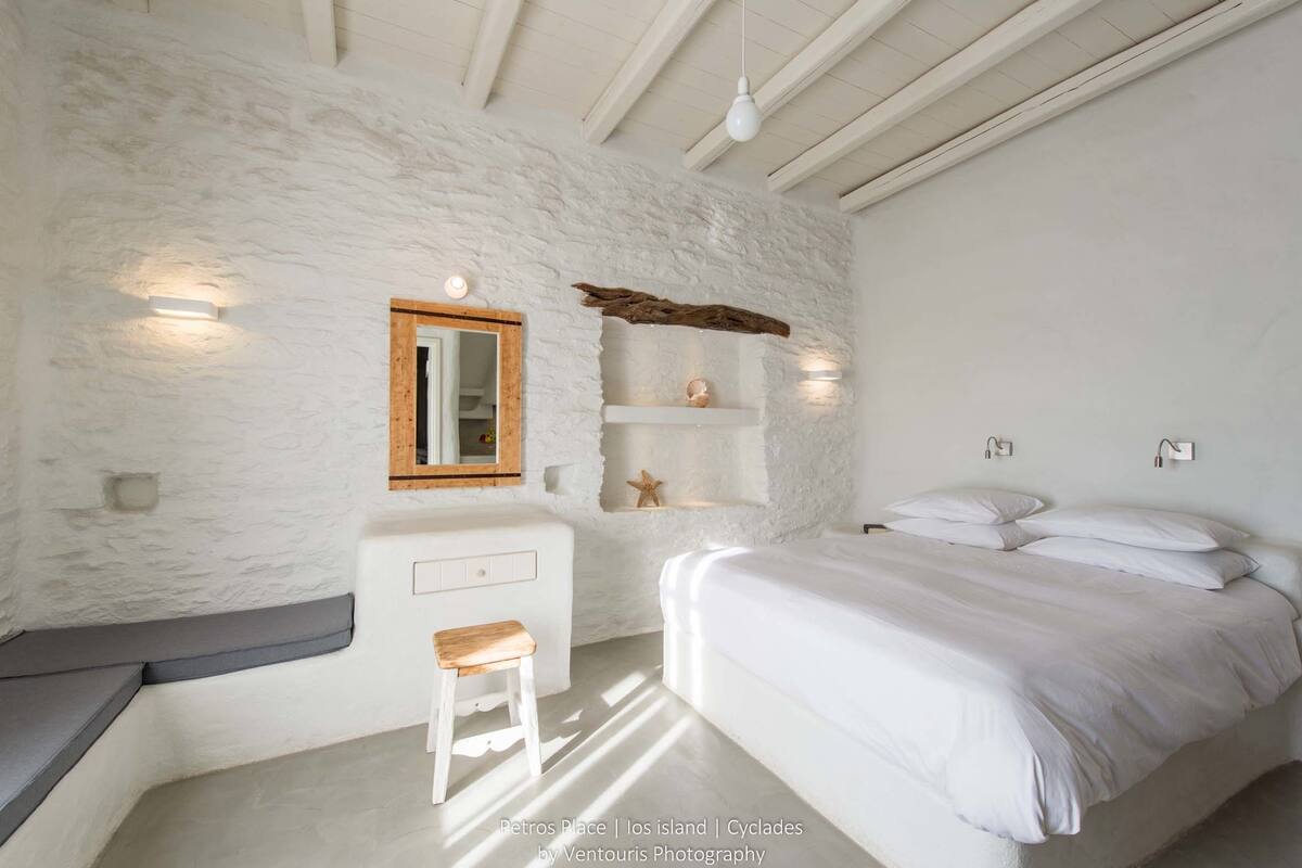 Quarto com parede de pedra branca, um banco de madeira, cama com roupa de cama branca, espelho com moldura de madeira e prateleira com objetos decorativos.