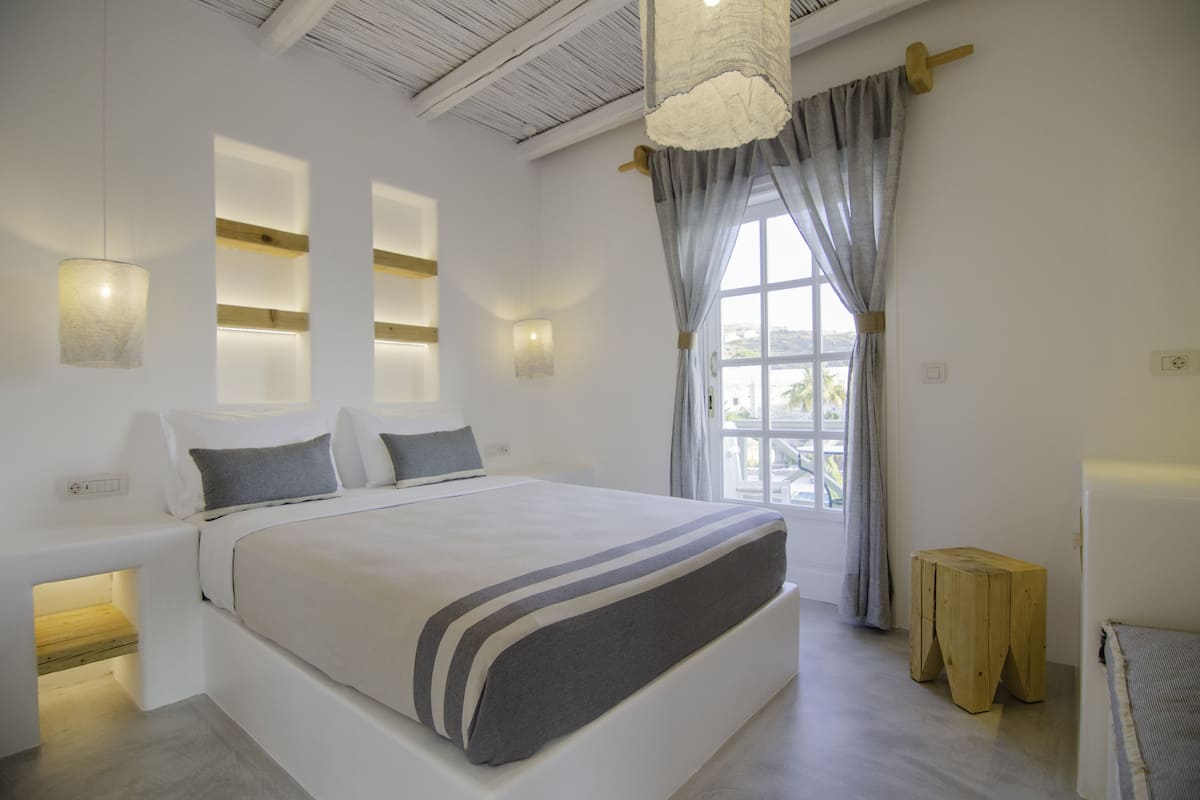 Quarto moderno com cama grande, abajures, um banquinho de madeira e vista para o exterior.
