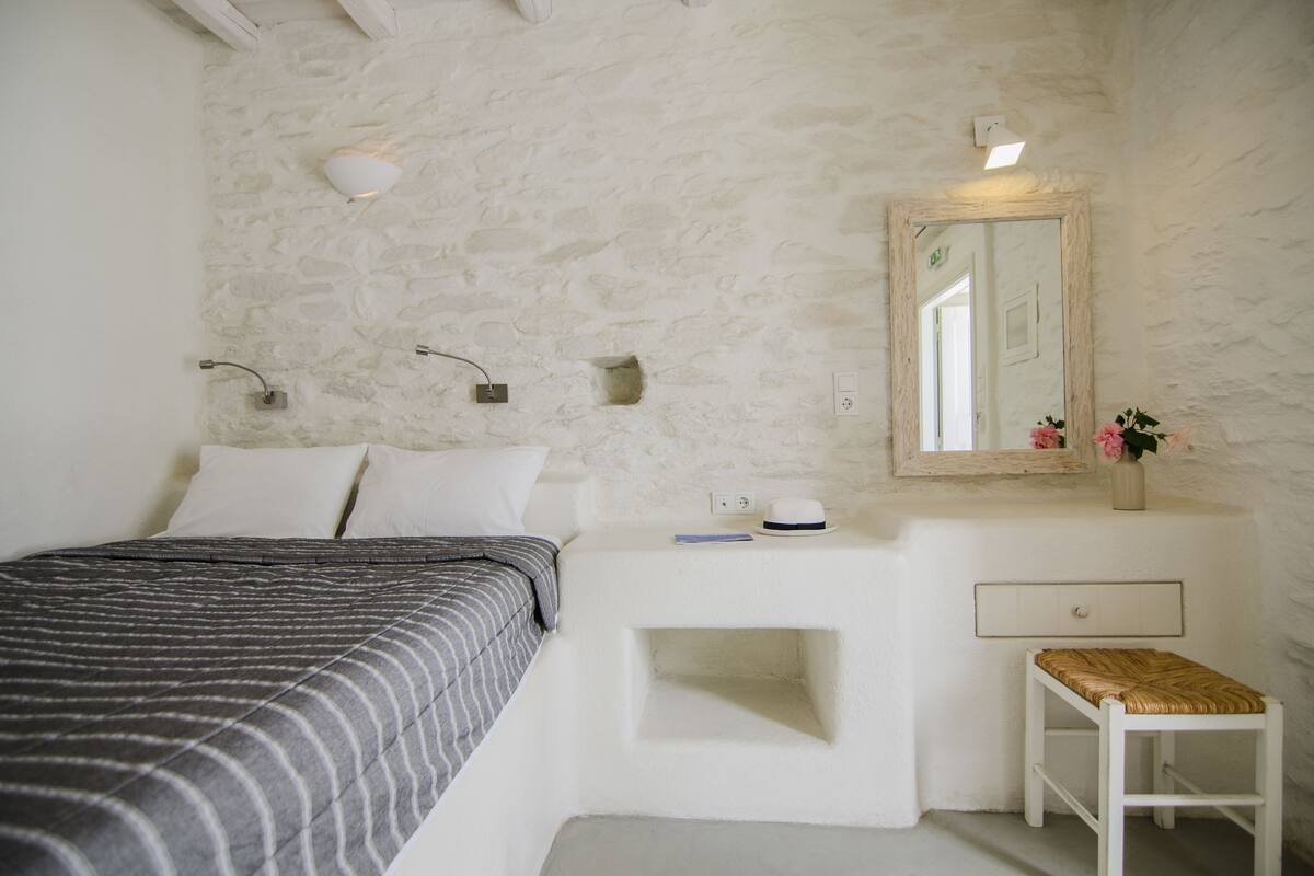 Quarto com cama, mesa de cabeceira, espelho e um banquinho.