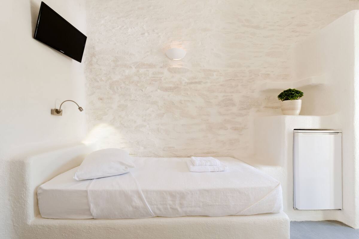 Quarto minimalista com uma cama branca, TV fixada na parede, luminária embutida, uma pequena planta em vaso e mini-geladeira integrada.
