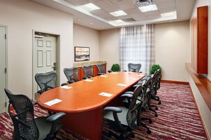 Uma sala de conferências com uma mesa comprida, várias cadeiras, uma pequena planta e um quadro na parede.
