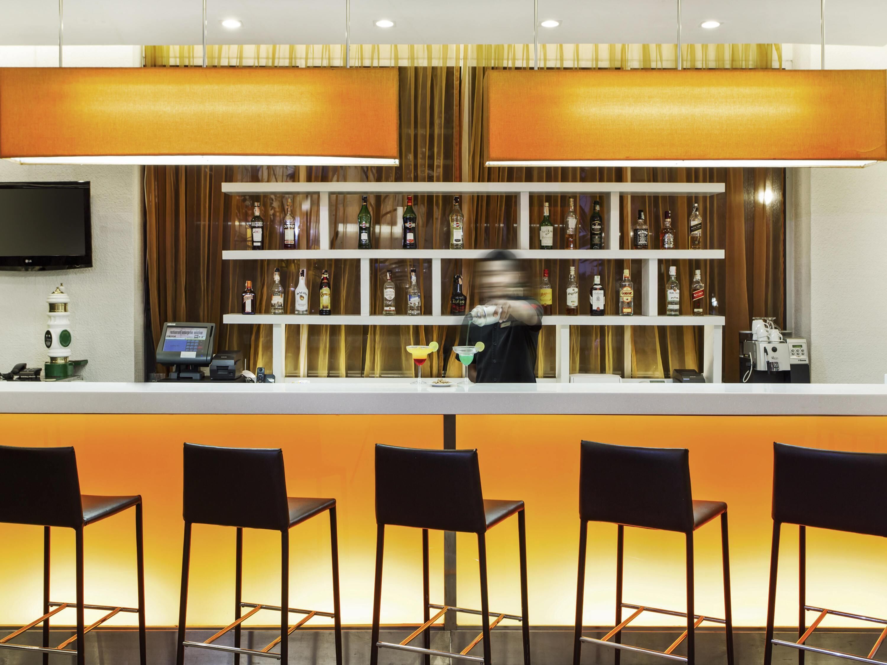 Uma área de bar com um bartender preparando bebidas, prateleiras com garrafas de bebidas alcoólicas, um caixa e banquetas de bar.