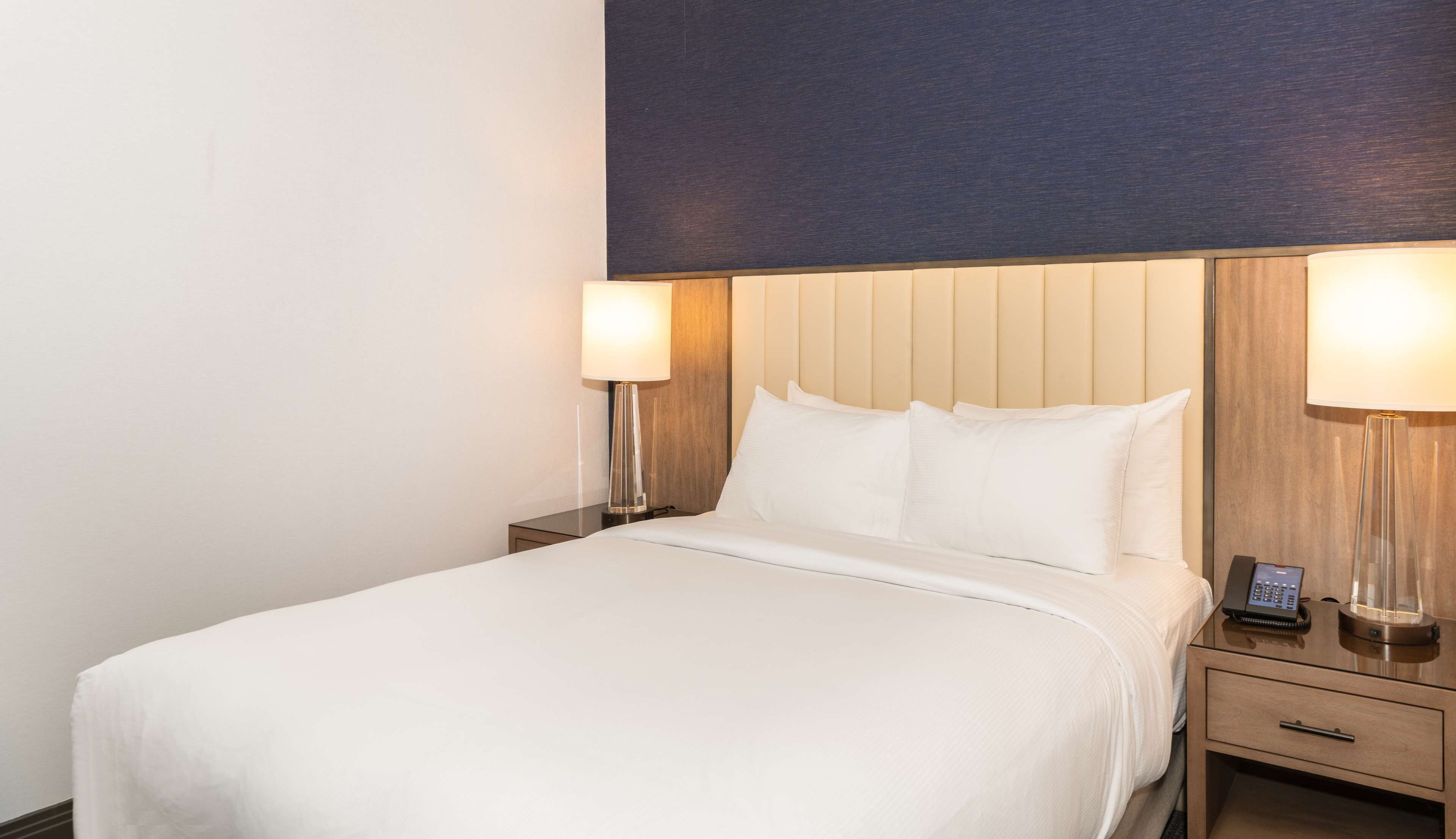 Quarto de hotel com uma cama, duas mesas de cabeceira e uma mesinha de apoio com telefone e luminária.