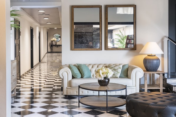 Um saguão de hotel com piso xadrez, um sofá branco com almofadas verdes, uma mesa de centro redonda e dois espelhos fixados na parede.