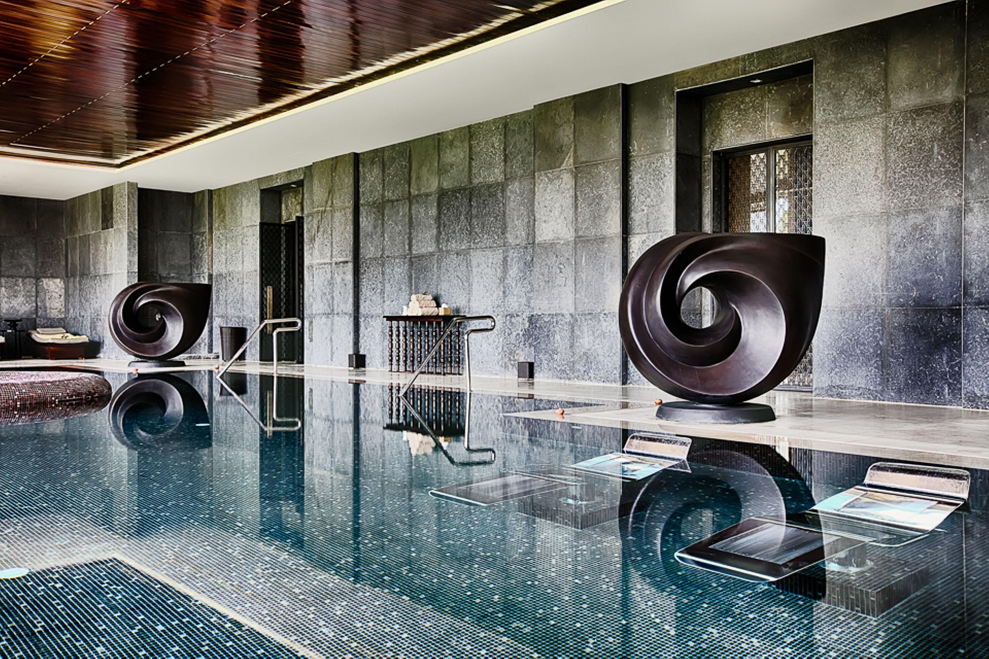 Piscina interior moderna com água cristalina, grandes esculturas abstratas e um painel de parede de pedra.