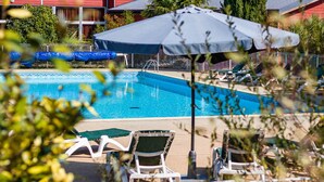 Piscina com espreguiçadeiras e guarda-sóis, cercada por uma vedação e vegetação.