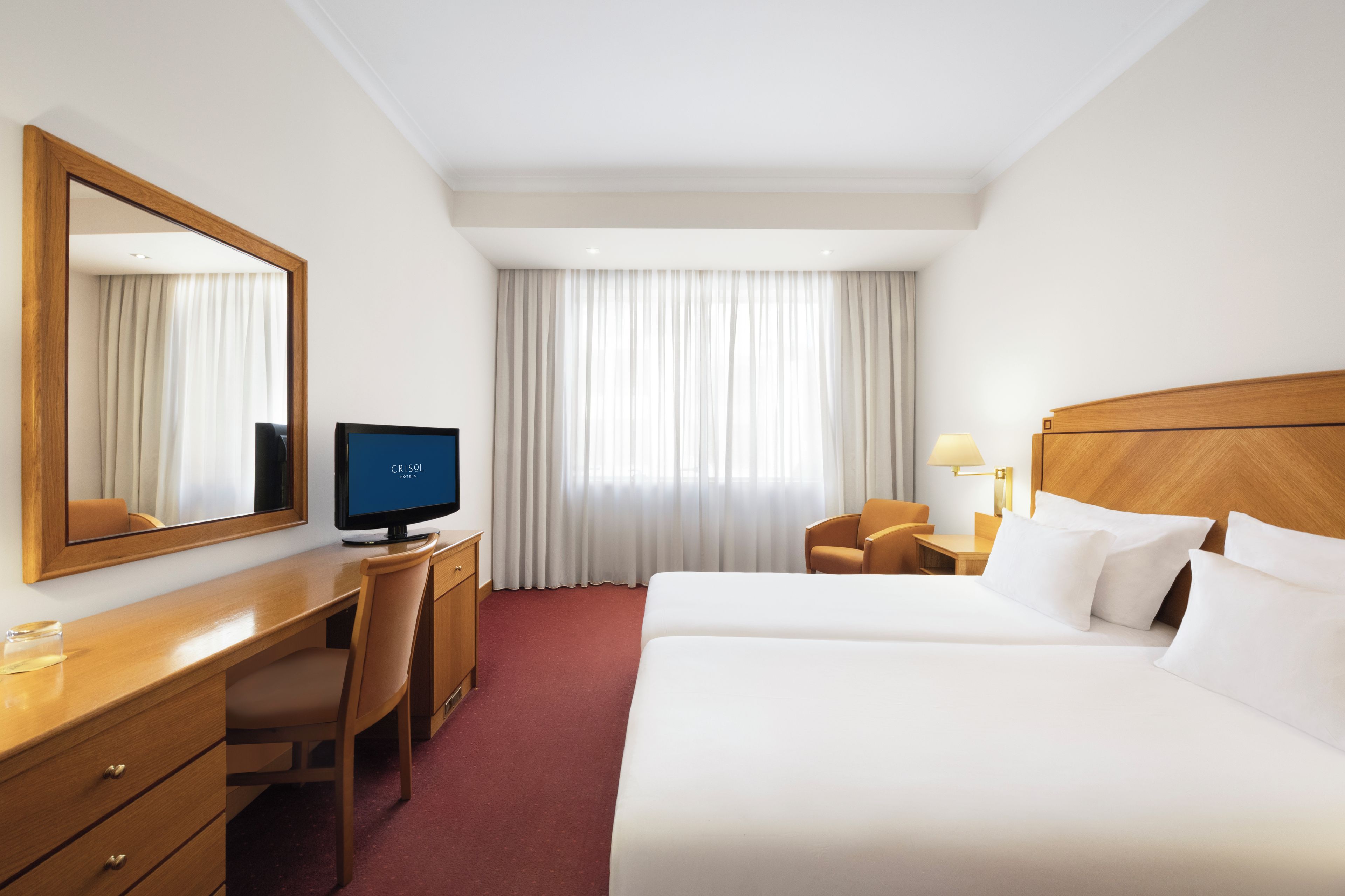 Quarto de hotel com cama, escrivaninha com computador, cadeira, abajur e um espelho grande.