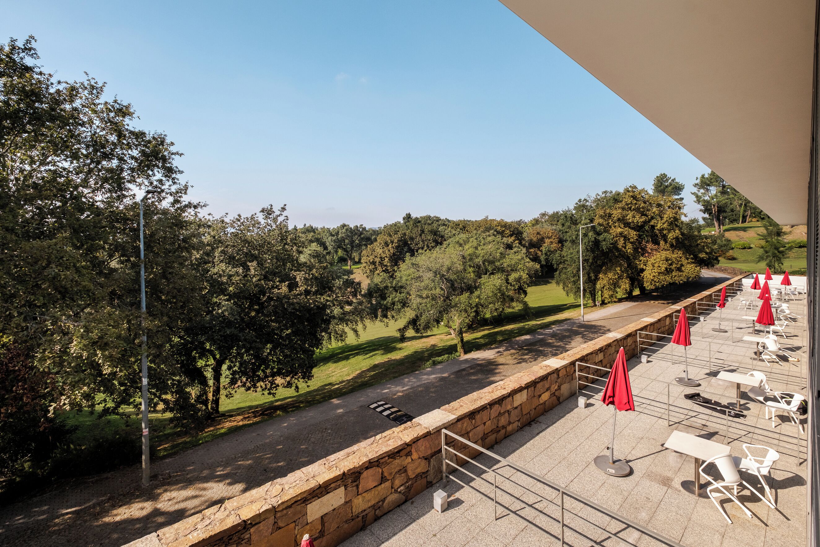 Varanda com grades de pedra com vista para a rua e vegetação, mobiliário de exterior com guarda-sóis vermelhos e céu limpo.