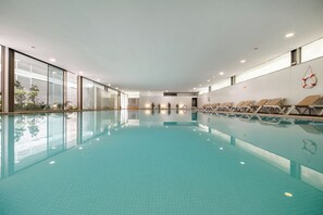 Uma grande piscina interior com água azul cristalina, espreguiçadeiras ao lado direito e um boi de salva-vidas na parede.