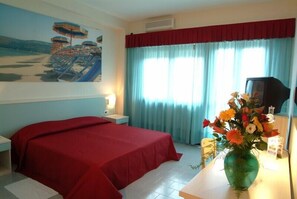 Quarto de hotel com uma cama, uma escrivaninha com um vaso de flores, uma televisão e um quadro com uma paisagem de praia.