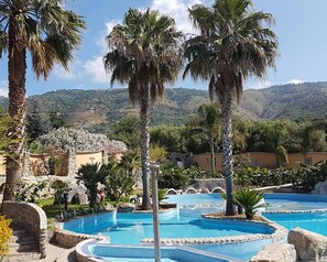 Área de piscina com palmeiras, arquitetura em pedra e montanhas ao fundo.