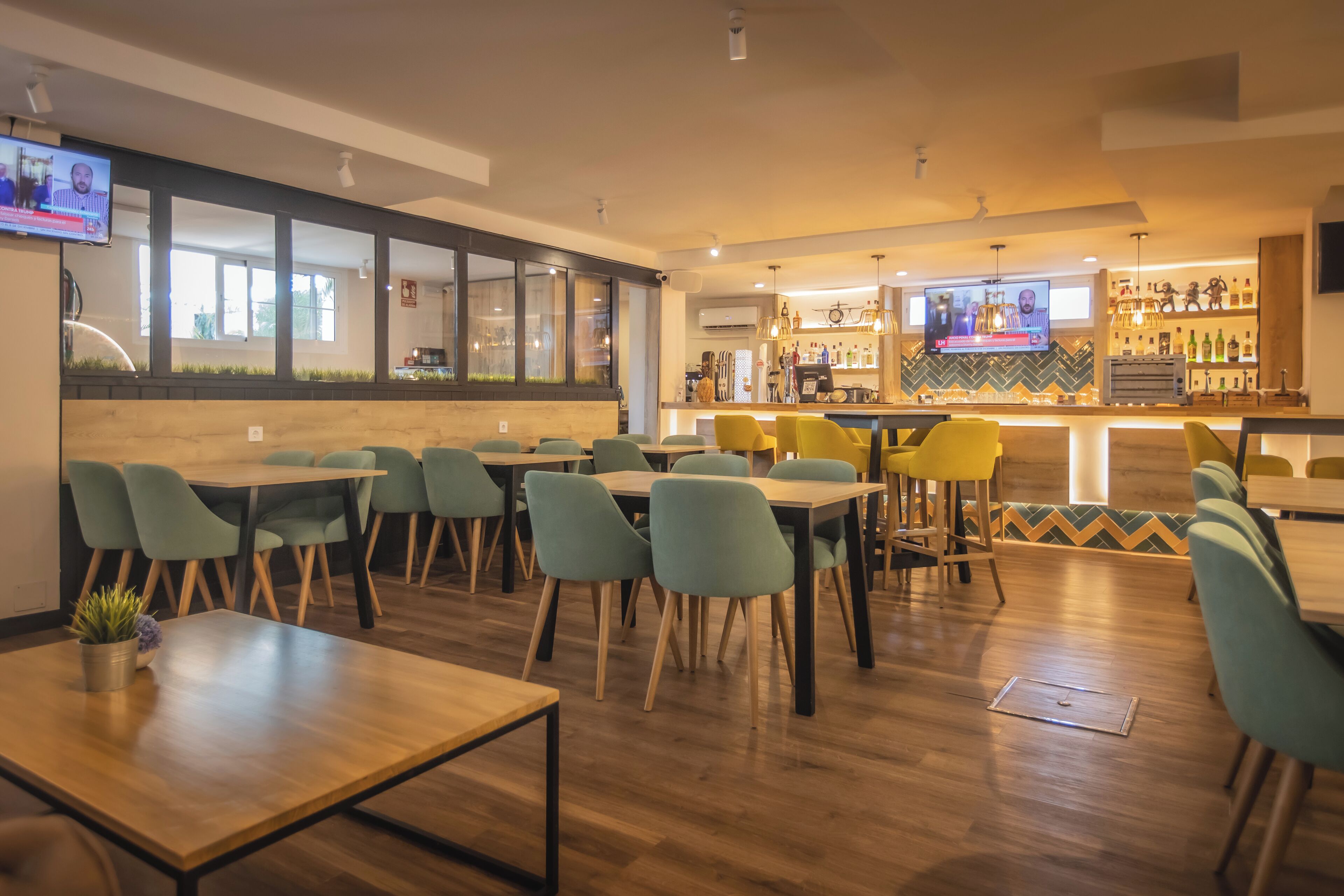 Uma área de bar moderna com chão de madeira, cadeiras verdes e amarelas, e um balcão com uma tela de televisão.