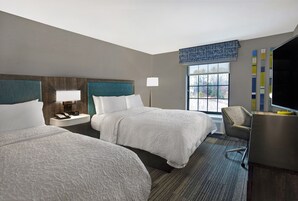 Quarto de hotel com duas camas, uma escrivaninha, uma cadeira, uma luminária e uma janela com vista.