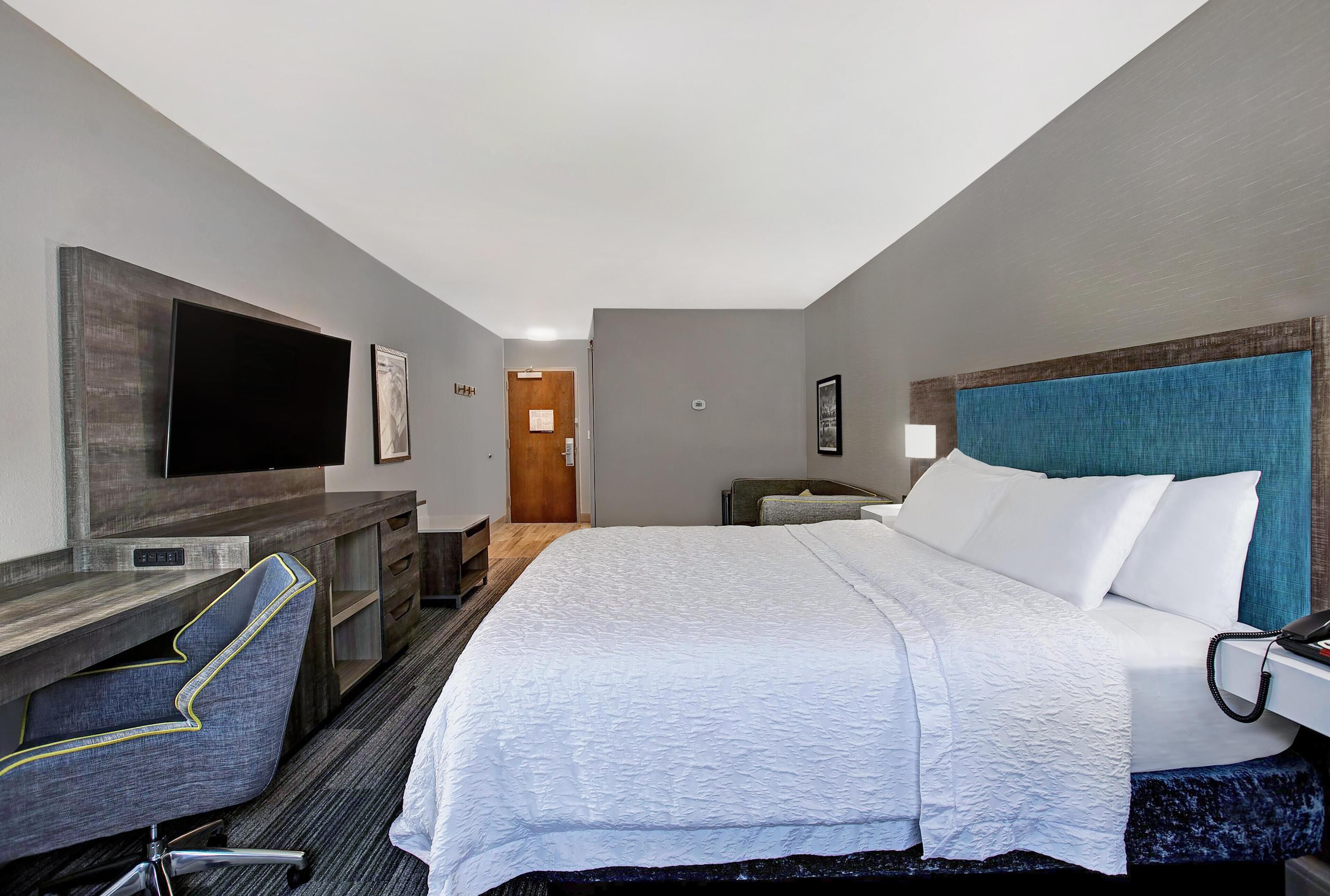 Quarto de hotel com uma cama grande, uma escrivaninha com cadeira, uma televisão de tela plana e uma porta de madeira.