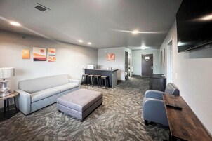 Quarto de hotel moderno com sofá, banquinho, área de bar e televisão de tela plana.