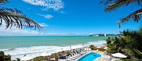 Área de piscina com espreguiçadeiras e guarda-sóis, com vista para uma praia e oceano.
