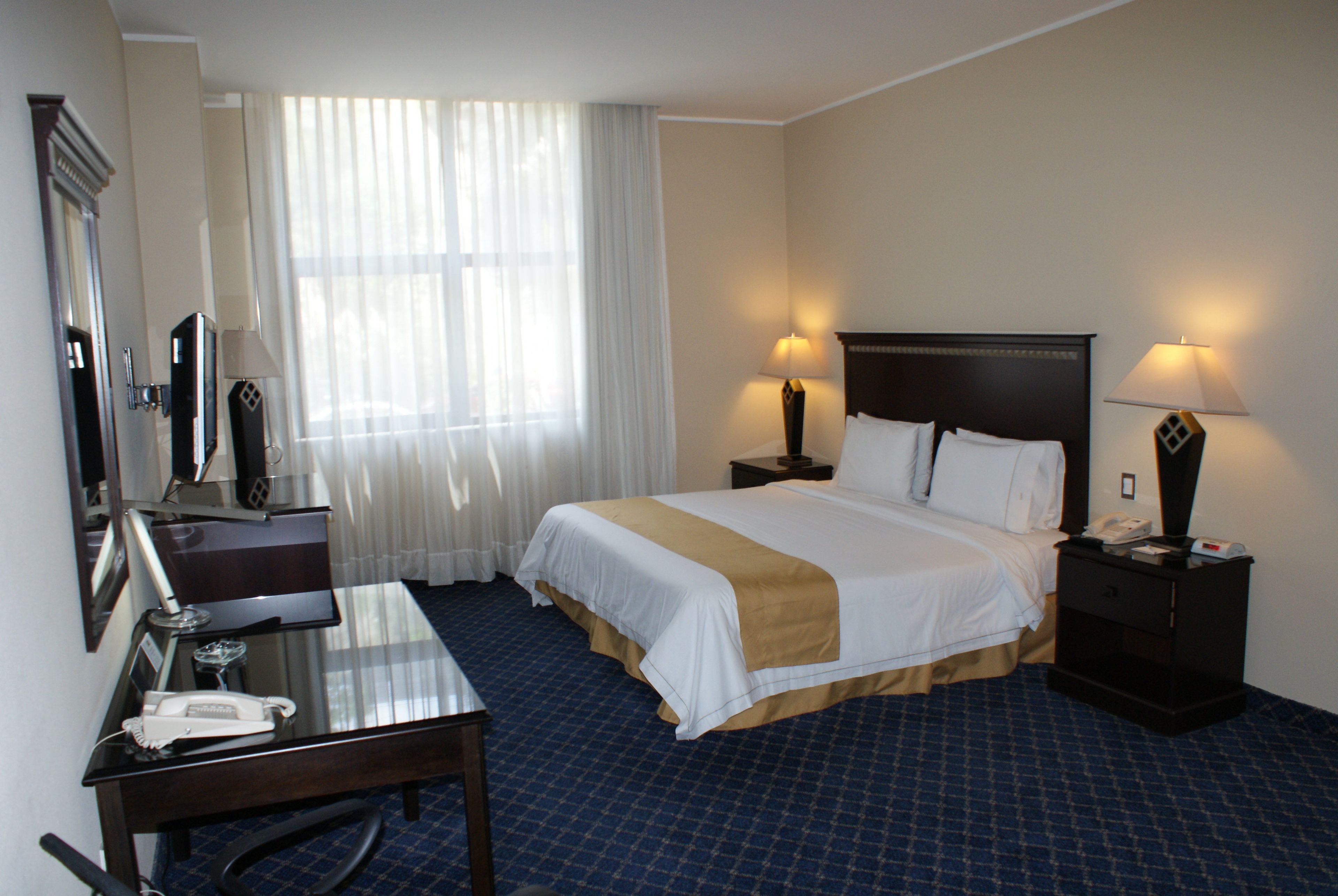 Quarto de hotel com cama, duas luminárias de cabeceira, escrivaninha com telefone e janela com cortinas.