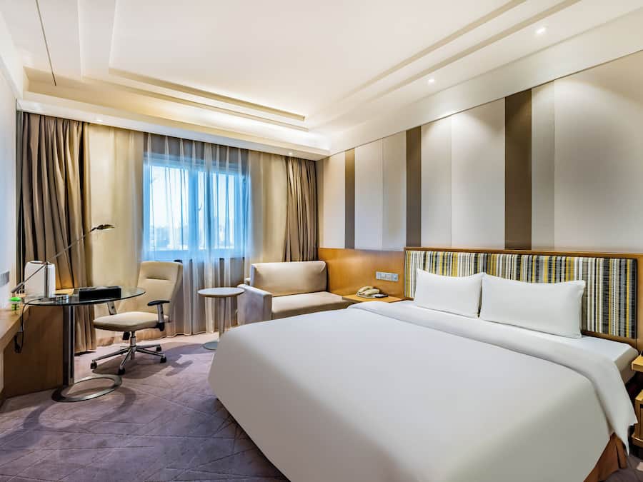 Quarto de hotel com uma cama grande, uma escrivaninha iluminada por um abajur, uma cadeira e uma mesinha.