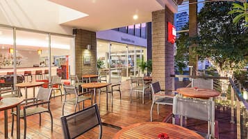 Uma área de café ao ar livre com mesas e cadeiras, um bar ao fundo e um edifício com uma placa.