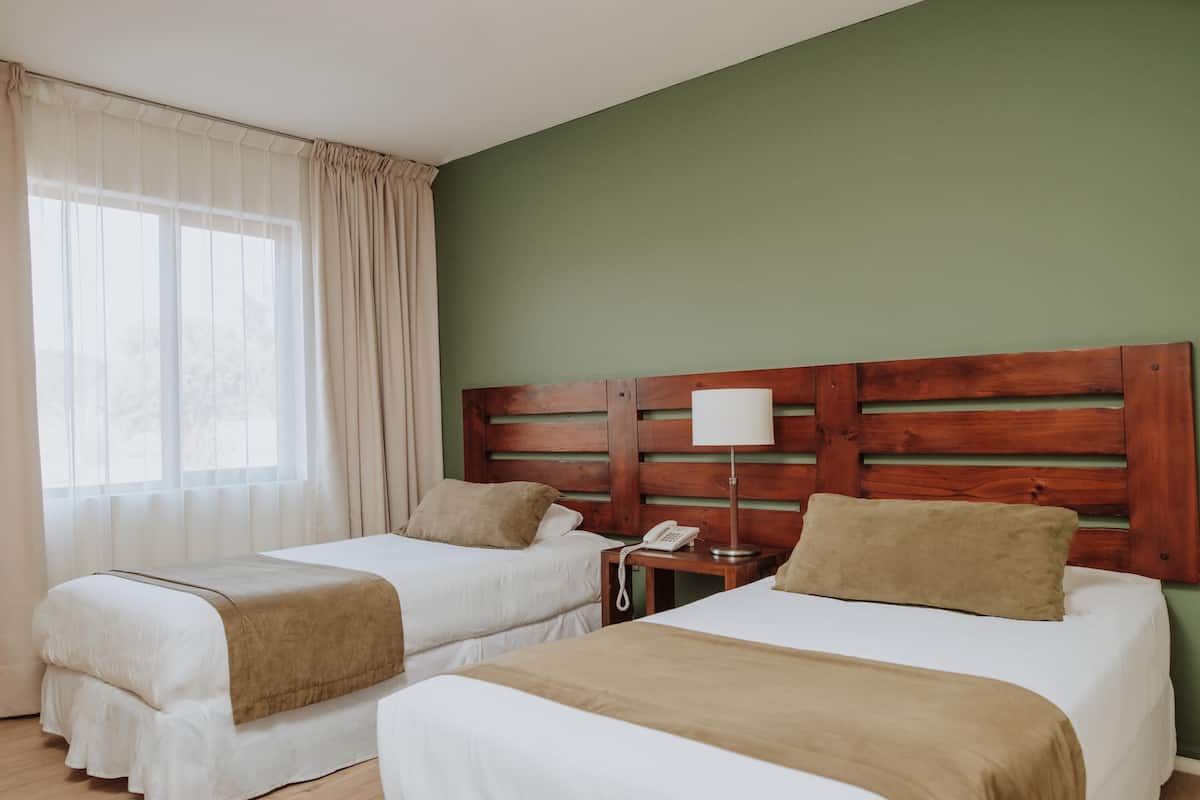 Quarto de hotel com duas camas, cabeceira de madeira, mesa de cabeceira com abajur e janela com cortinas.