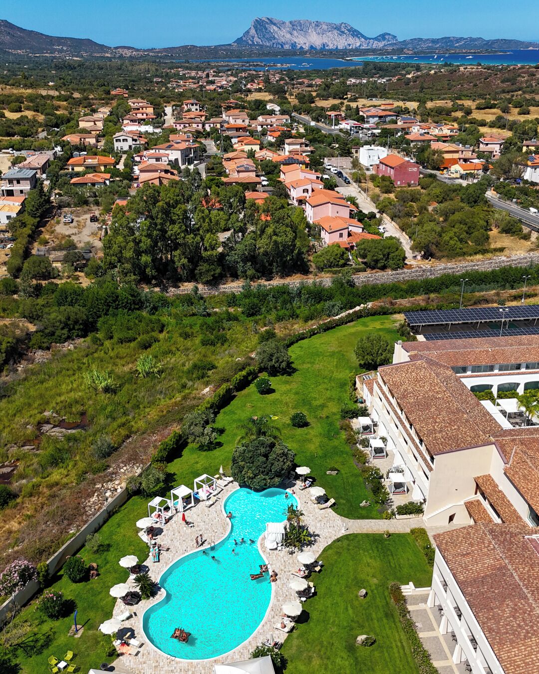 Vista aérea de uma área residencial com piscina, cadeiras de descanso e guarda-sóis, um edifício grande com telhado de telhas vermelhas e montanhas ao fundo.