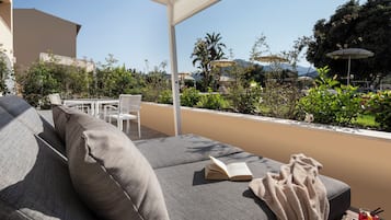 Varanda com sofá cinzento, uma mesa e cadeiras, cercada por vegetação e vista para edifícios.