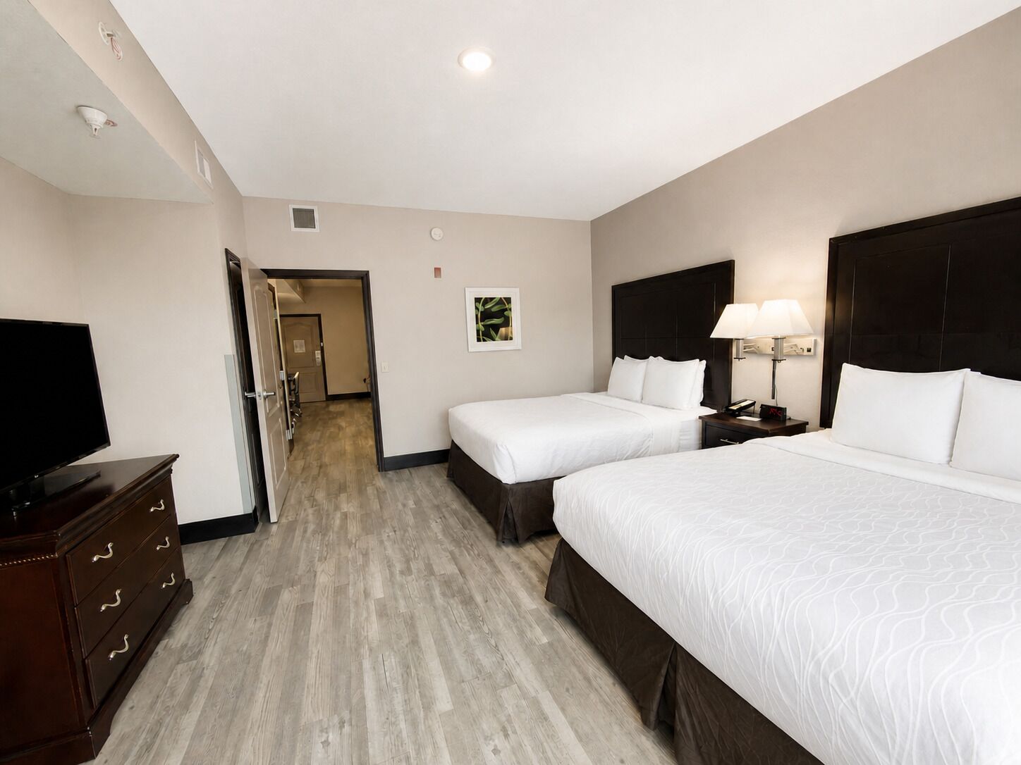 Quarto de hotel com duas camas, televisão, penteadeira e corredor que dá para outro quarto.