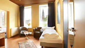 Quarto de hotel com cama, escrivaninha, cadeira, mesa pequena com um vaso e janelas amplas com cortinas.