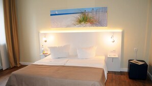 Uma cama bem arrumada com um cobre-leito bege, duas mesas de cabeceira com abajures, e uma pintura de uma paisagem praiana sobre a cabeceira.