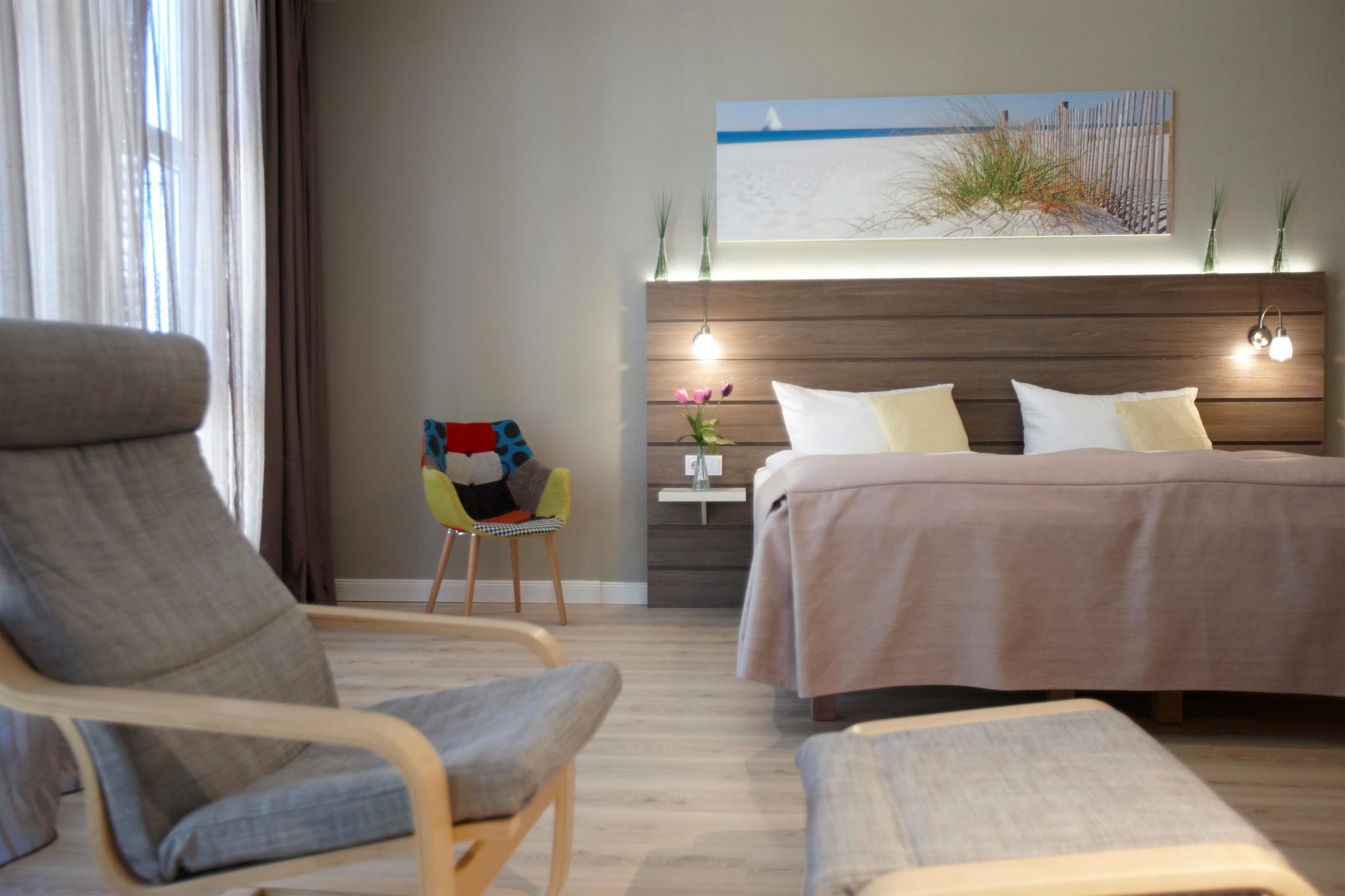Quarto de hotel moderno com uma cama grande, área de estar com uma cadeira, uma mesinha e uma obra de arte na parede retratando uma cena de praia.