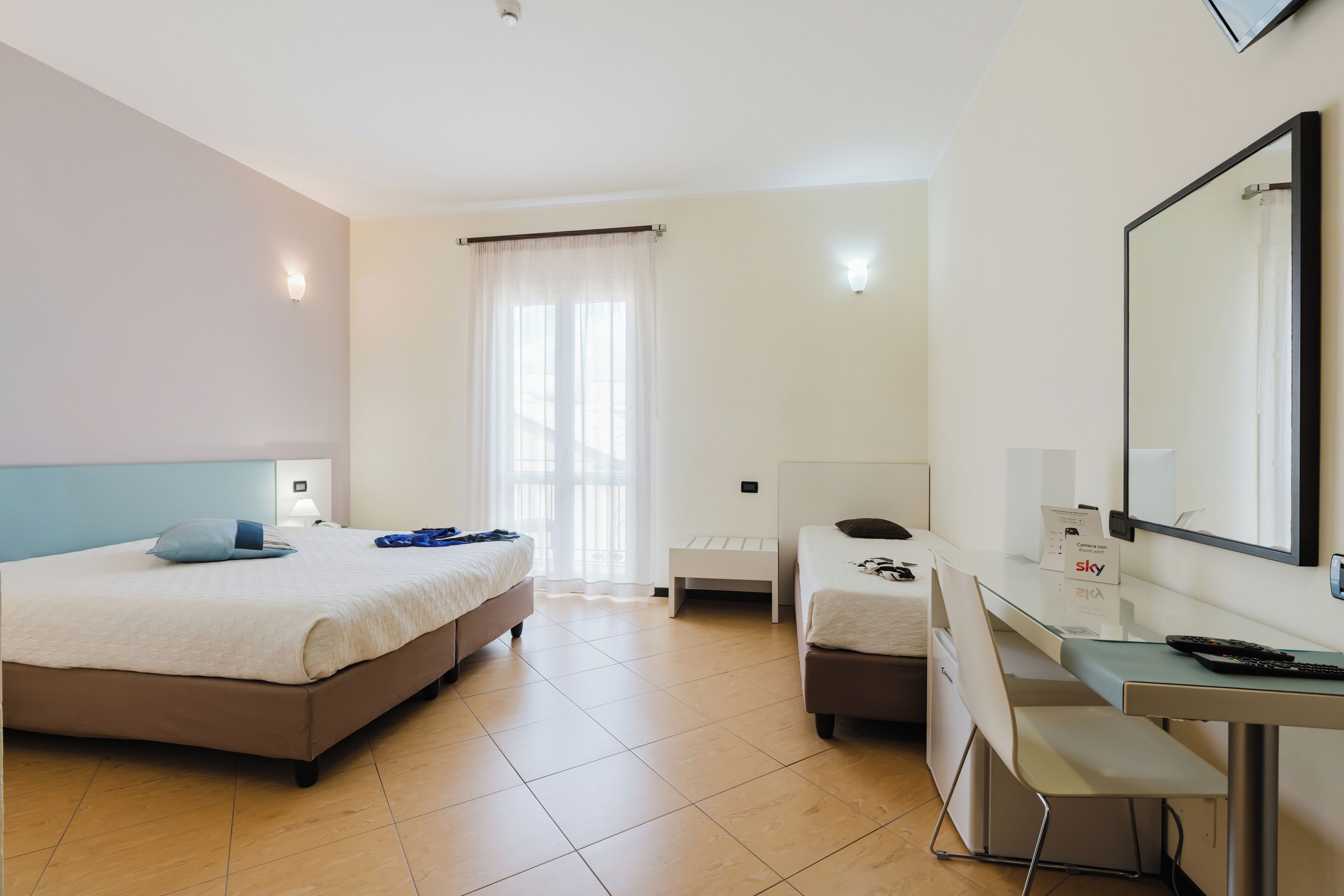 Quarto de hotel com uma cama, escrivaninha, cadeira, espelho, janela com cortinas e mesa de cabeceira.