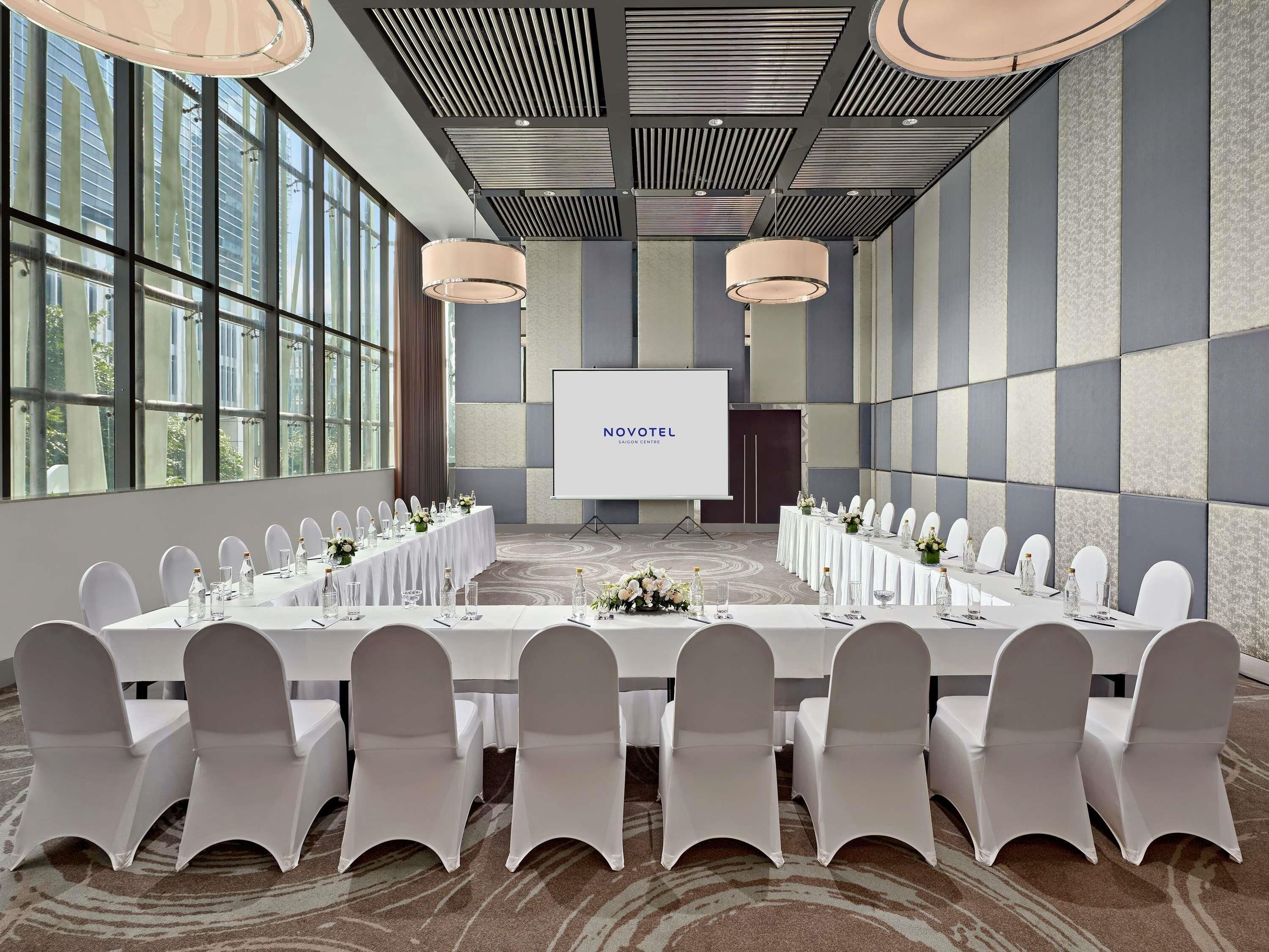 Uma sala de conferências com uma mesa comprida, cadeiras brancas e uma tela de projeção exibindo 'NOVOTEL'.