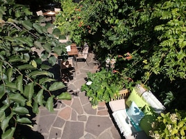 Um espaço ao ar livre com piso de pedra, uma mesa e cadeiras de madeira, cercado por vegetação exuberante e plantas em vasos.