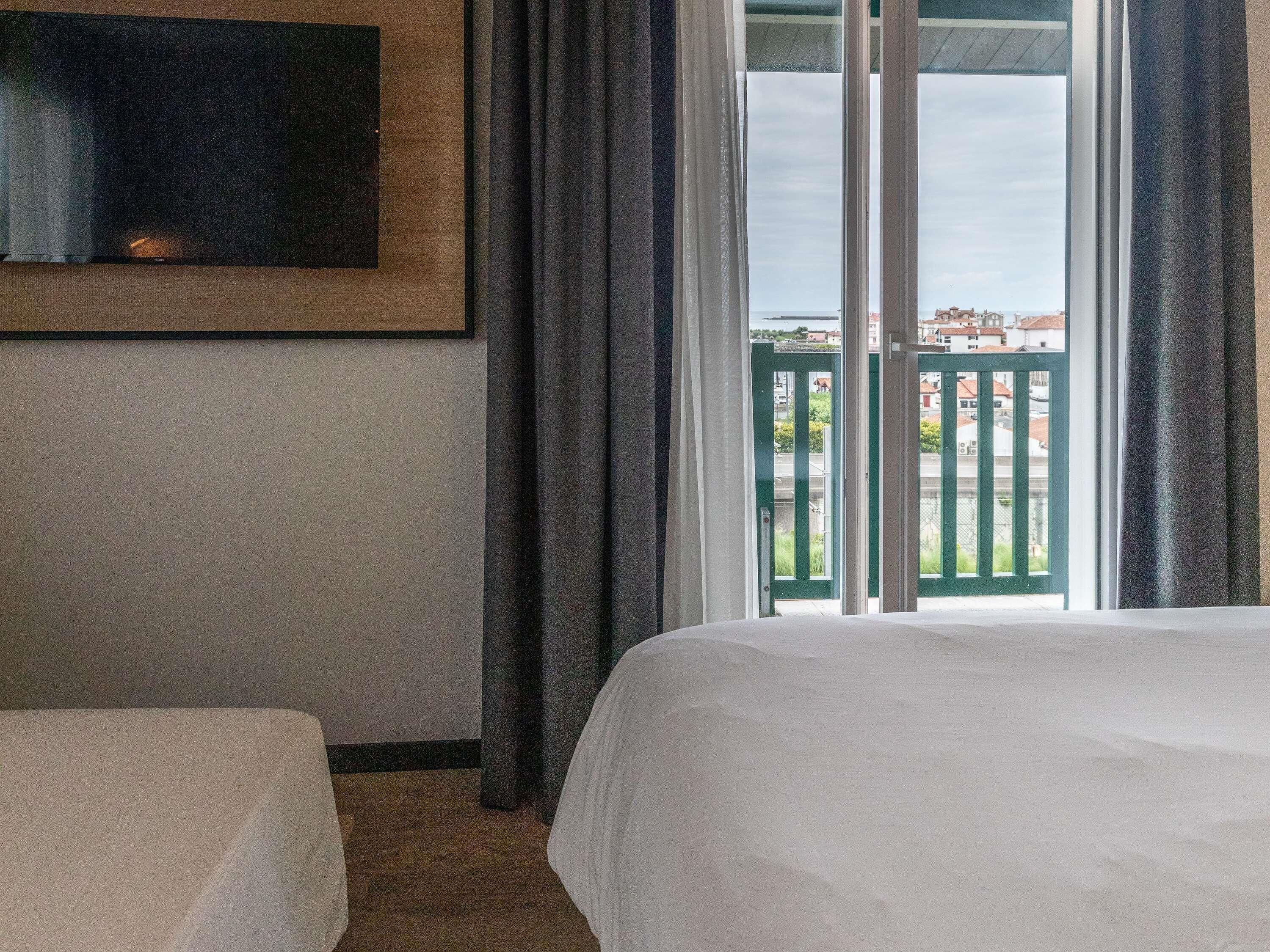 Quarto de hotel com uma cama, televisão na parede e vista para uma varanda com guarda-corrimão verde e edifícios ao fundo.