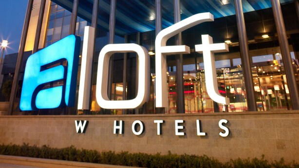 Entrada de hotel com design moderno, destacando uma placa 'aLoft' e fachada de vidro.
