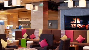 Uma área de estar moderna com sofás, poltronas e uma mesa de centro. Há uma lareira acesa e uma placa indicando uma saída.