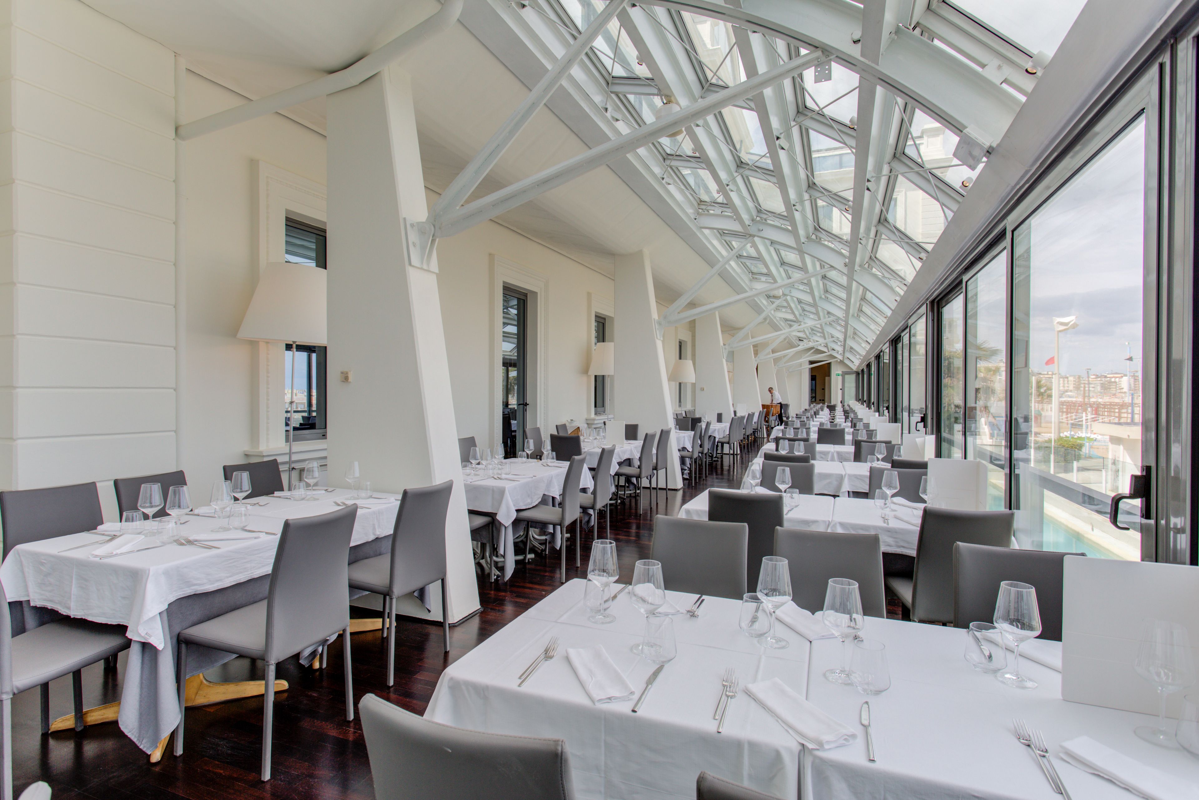 Um restaurante espaçoso com pé direito alto e janelas grandes, com mesas e cadeiras organizadas.