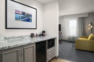 Quarto de hotel com bancada de mármore, televisão, sofá, cadeira e um quadro na parede.