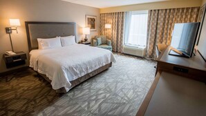 Quarto de hotel com uma cama grande, duas mesas de cabeceira, uma cadeira, uma televisão e uma janela com cortinas.