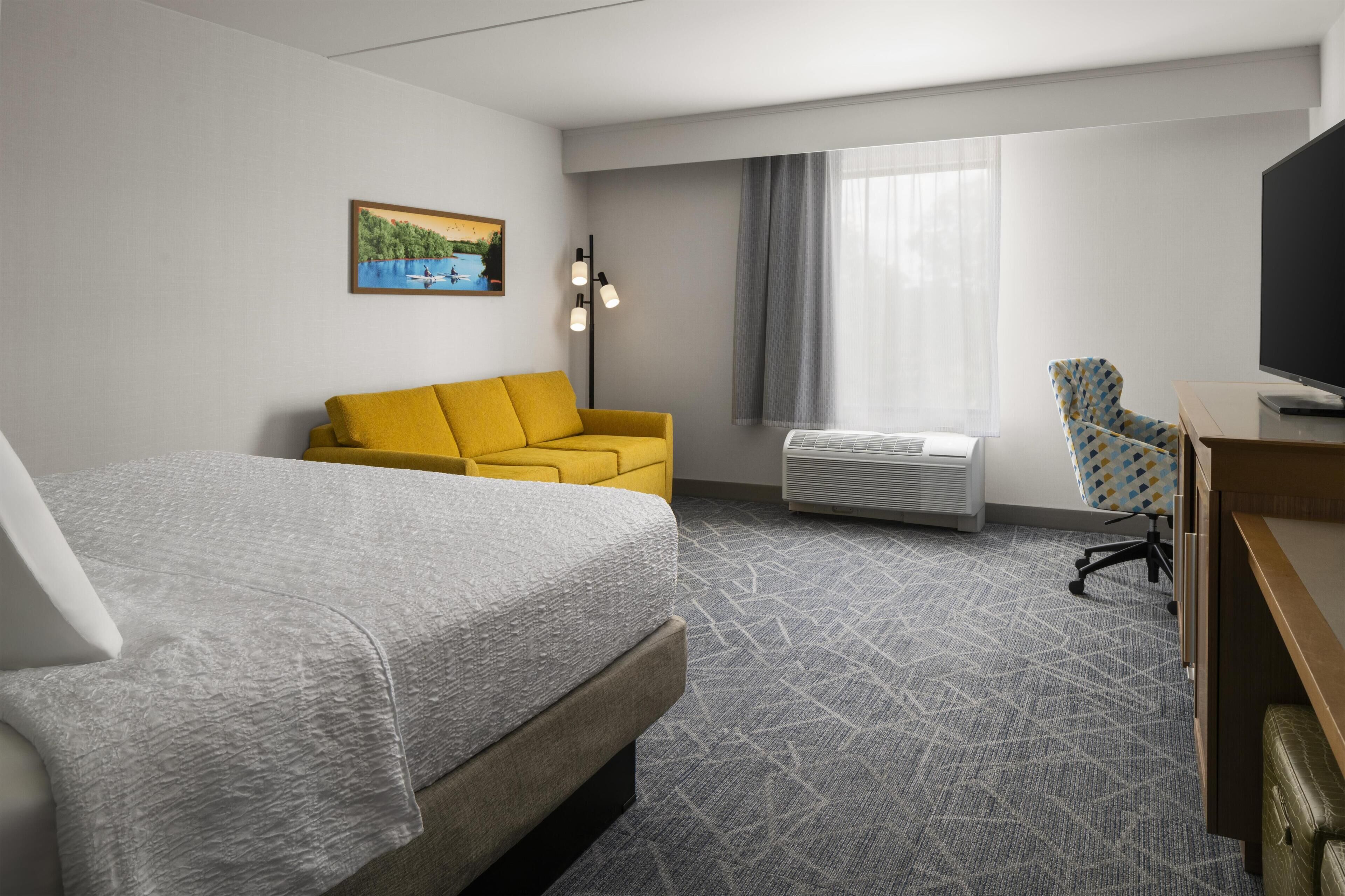 Quarto de hotel com uma cama, uma poltrona amarela, uma mesa de escritório com cadeira, uma televisão e um quadro na parede.