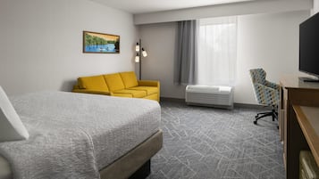 Quarto de hotel com uma cama, uma poltrona amarela, uma mesa de escritório com cadeira, uma televisão e um quadro na parede.