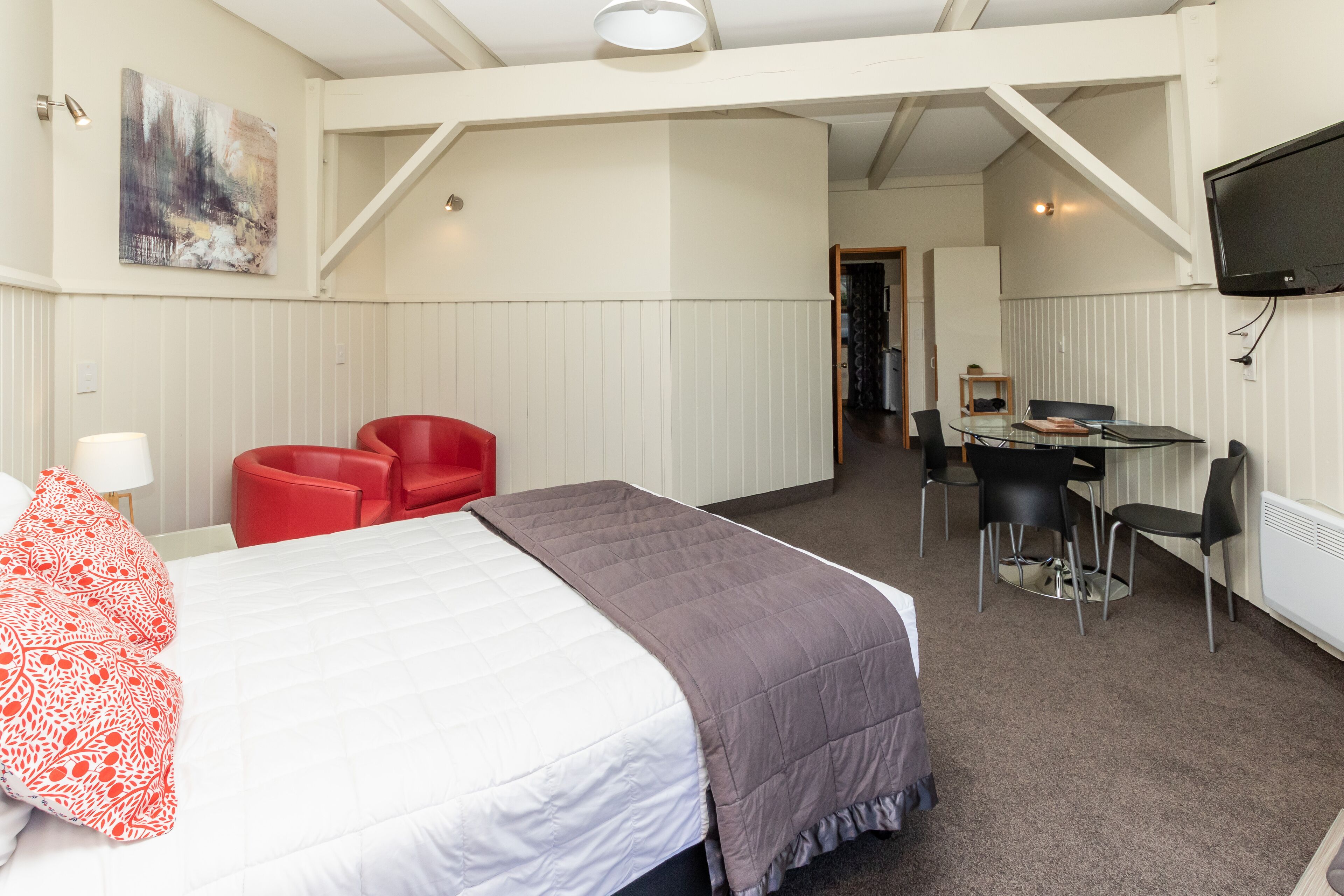 Quarto de hotel com cama, cadeiras vermelhas, escrivaninha e televisão.