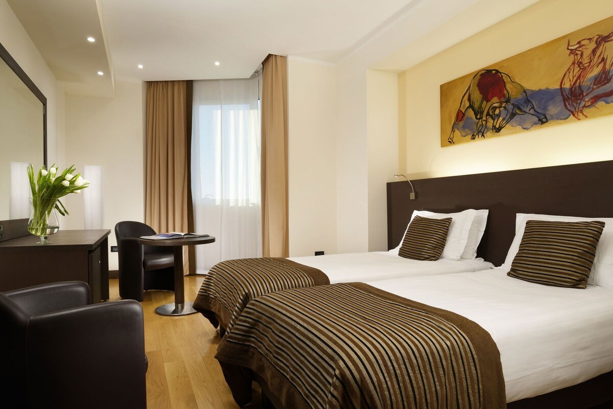 Quarto de hotel com duas camas, uma escrivaninha, uma cadeira e um quadro na parede.